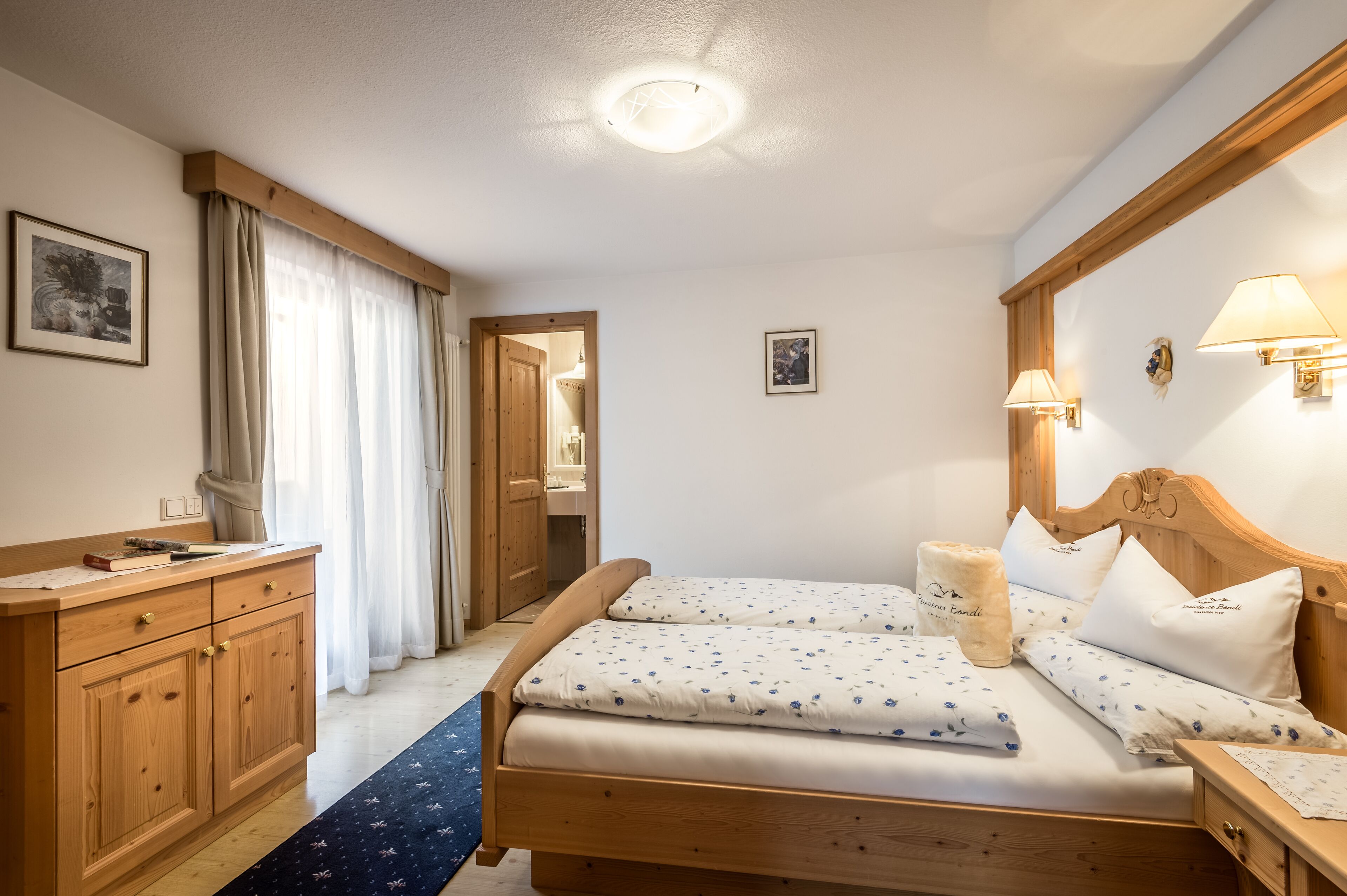 Quarto de hotel com cama de madeira, mesa de cabeceira, escrivaninha, cadeira, janela com cortinas, luz de teto e luminária na parede.