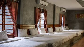 Quarto de hotel com quatro camas individuais alinhadas, cada uma com roupa de cama branca e várias almofadas. Há janelas amplas com cortinas vermelhas e um aparelho de ar condicionado fixado na parede.