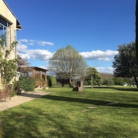 Um gramado bem cuidado com uma pérgola de madeira, estruturas de pedra e diversas árvores.