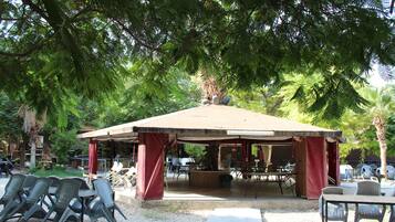 Um pavilhão ao ar livre com telhado e área de estar, rodeado por árvores e vegetação.