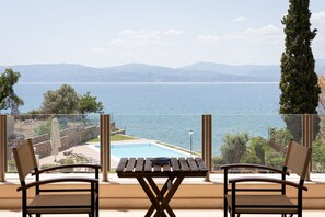 Varanda com duas cadeiras de vime e uma mesa de madeira, com vista para uma piscina e um corpo d'água com montanhas ao longe.