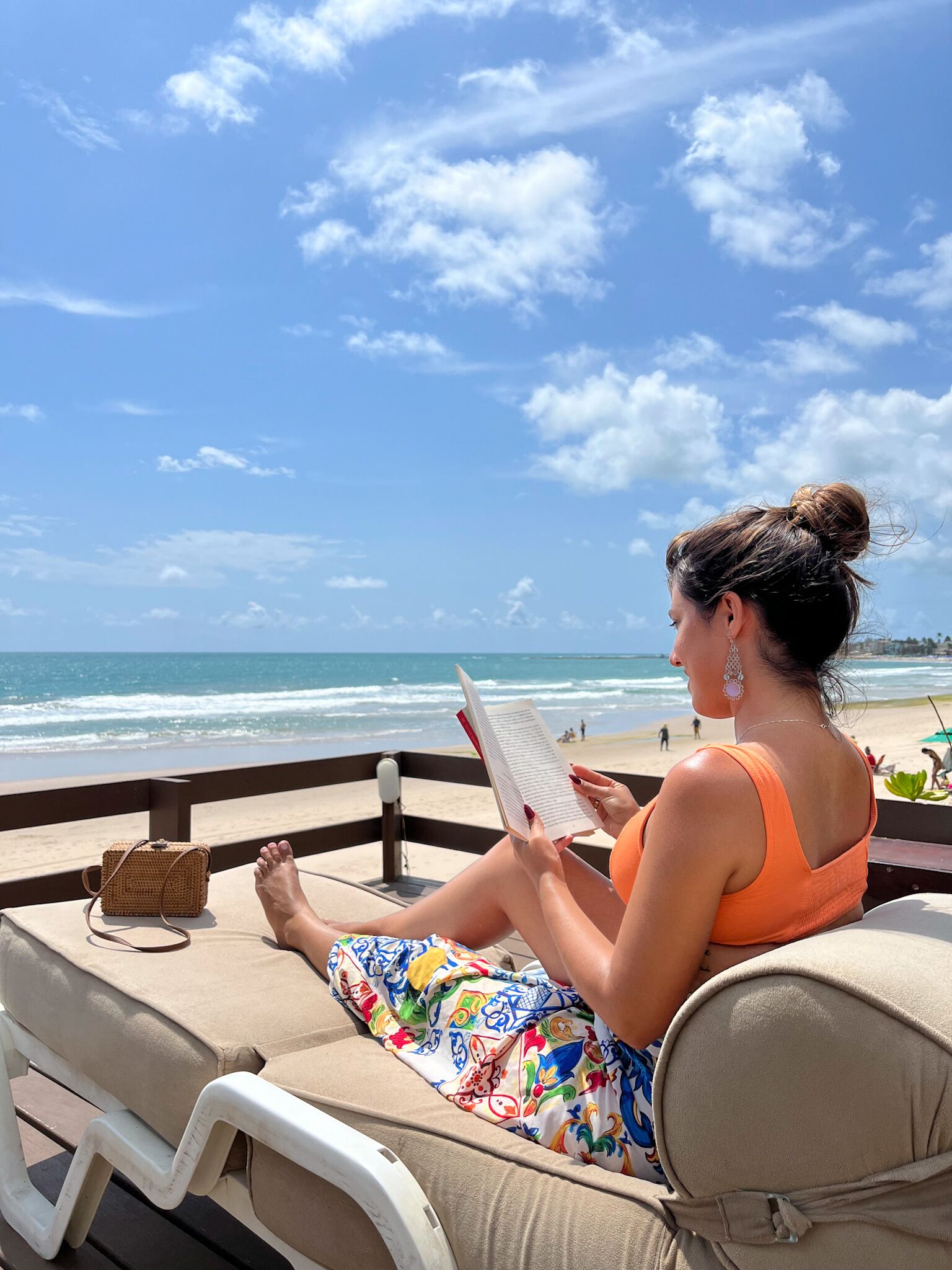 Uma pessoa sentada numa cadeira de praia lendo um livro, com vista para a praia e o oceano.