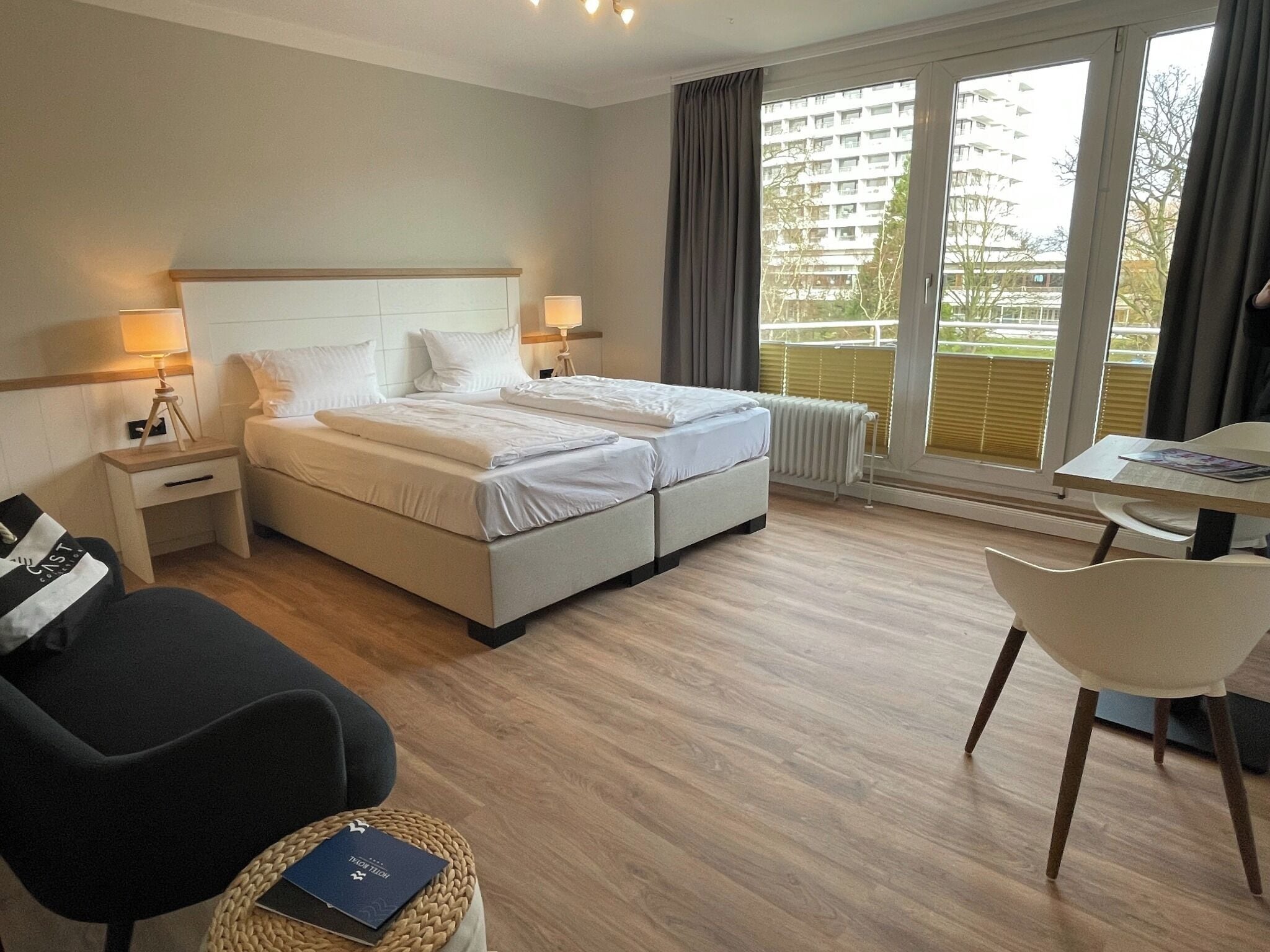 Quarto de hotel com cama, mesas de cabeceira, uma cadeira, uma pequena mesa e vista para o exterior.
