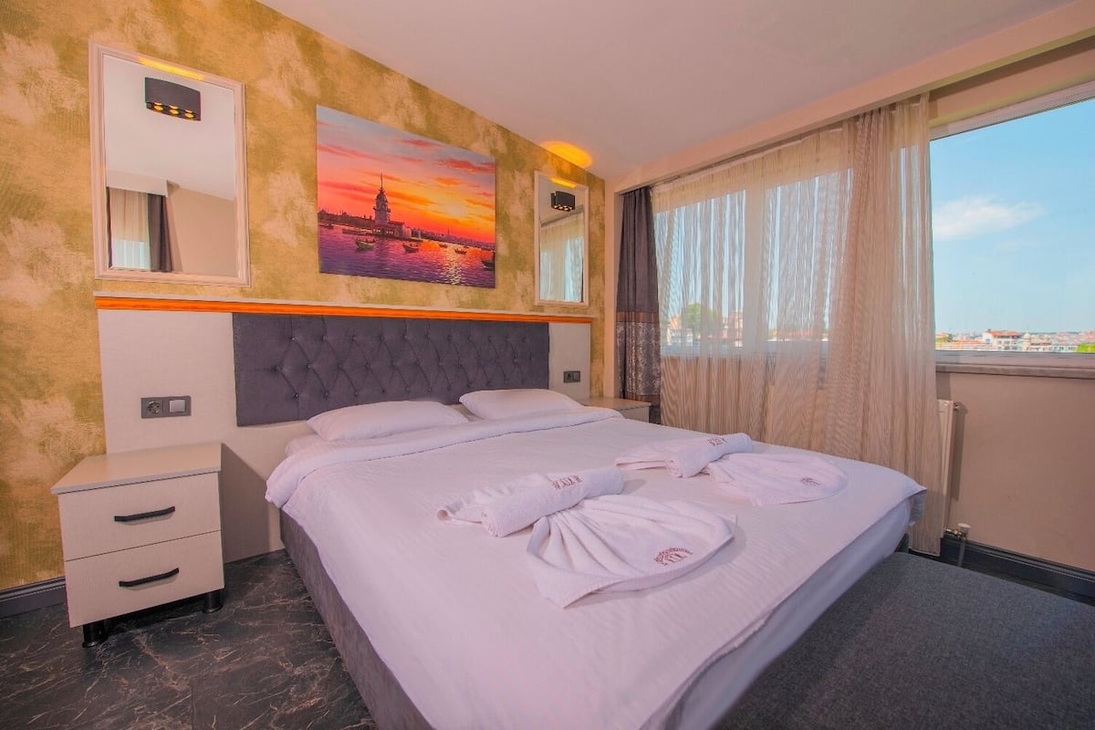 Quarto de hotel com cama, mesa de cabeceira, espelho, quadro e janela com cortinas.