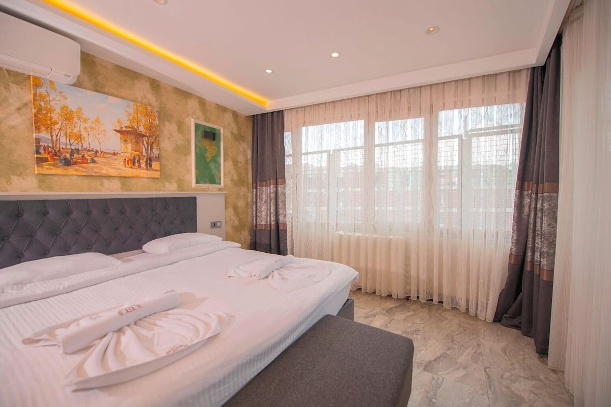 Quarto de hotel com cama grande, cabeceira, um quadro na parede e janela com cortinas.