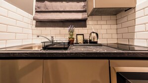 Uma cozinha com paredes revestidas de azulejos brancos, bancada de mármore preto, pia, janela com persiana marrom e um vaso com planta.