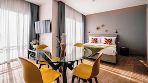 Quarto de hotel moderno com mesa de jantar de vidro, cadeiras amarelas, cama com manta verde e TV fixada na parede.