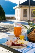 Uma mesa com uma refeição, incluindo um jarro de suco de laranja, uma xícara e um prato com queijos e carnes, ao fundo de uma paisagem montanhosa.