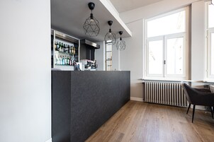 Uma área de bar moderna com balcão escuro, piso de madeira e luminárias pendentes geométricas.