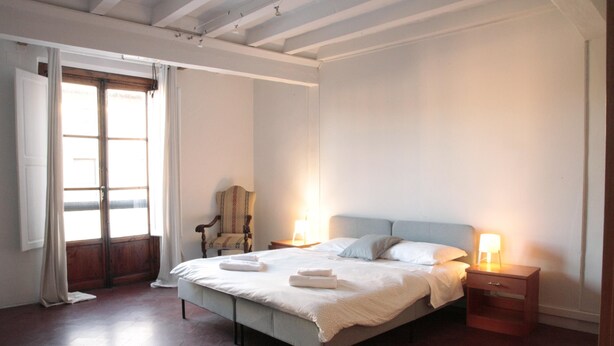 Quarto com cama grande, uma cadeira, mesa de cabeceira e janela com cortinas.