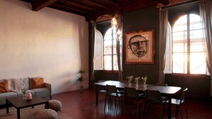 Uma sala de estar ampla com sofá, mesa de centro e mesa de jantar. Há uma janela grande com cortinas, um quadro na parede e um vaso de planta.