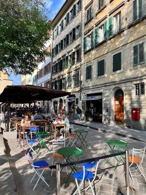 Um café ao ar livre com mesas e cadeiras, rodeado por edifícios históricos com persianas verdes.