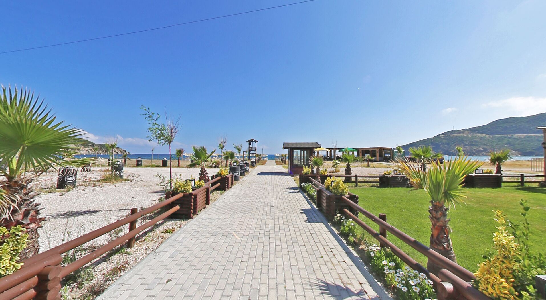 Uma trilha pavimentada leva a uma área de praia com palmeiras e um gazebo.