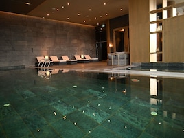Piscina interior moderna com espreguiçadeiras e área de recepção em madeira.