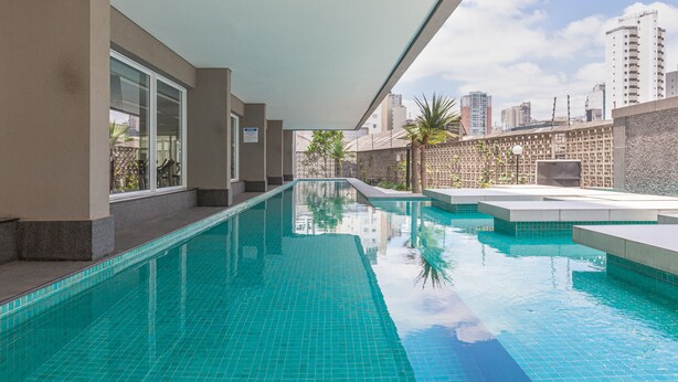 Uma piscina com água azul cristalina, rodeada por um edifício moderno com grandes janelas e vista para a paisagem urbana ao fundo.