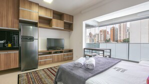 Quarto de hotel moderno com uma cama grande, televisão, frigobar e varanda com vista para a cidade.