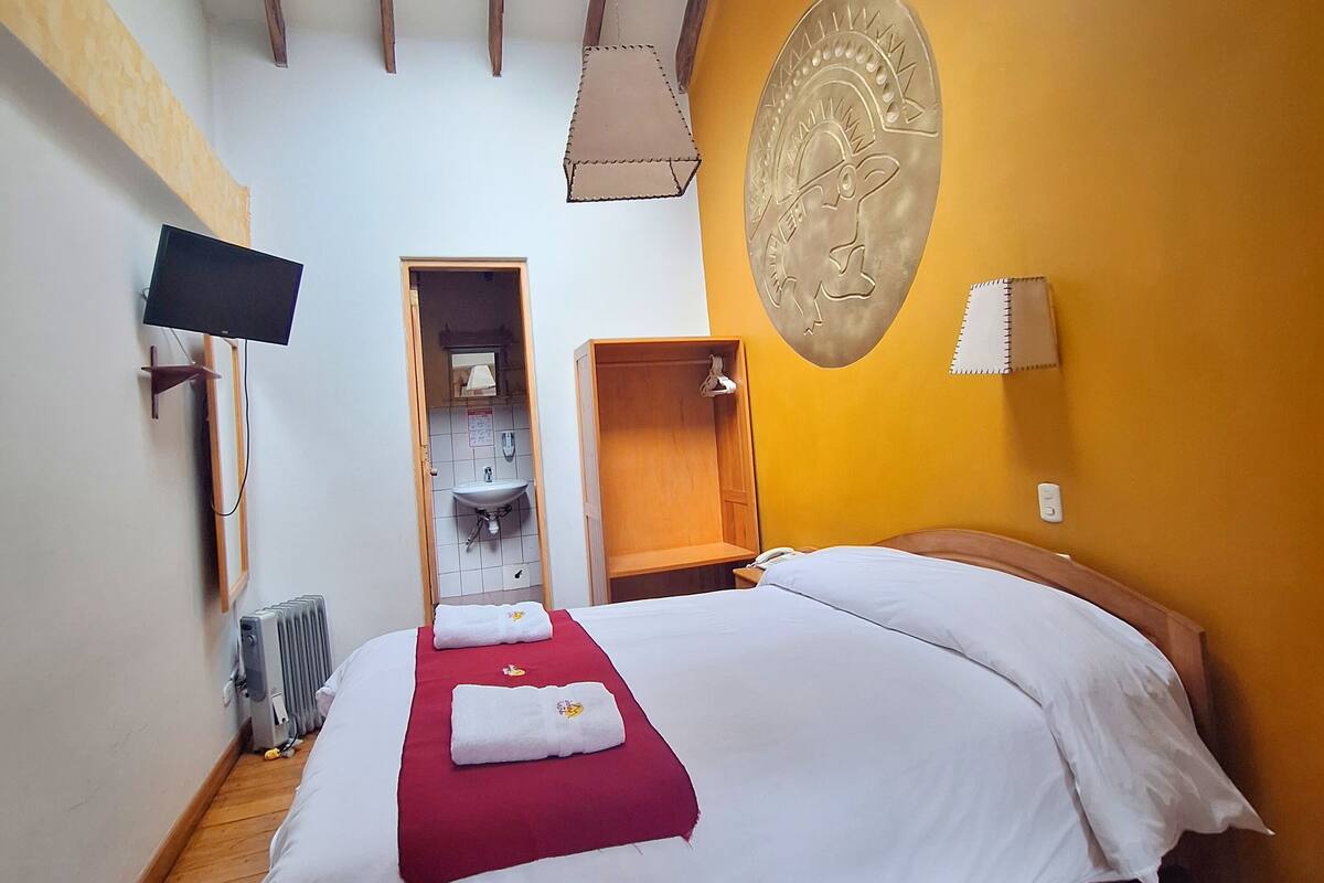 Quarto com cama grande, vigas de madeira, claraboia, ornamento circular na parede, mesa de cabeceira e radiador.