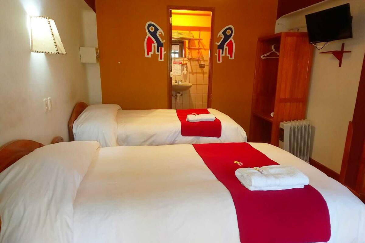 Quarto de hotel com duas camas, cada uma com lençóis brancos e um cobertor vermelho. Há um guarda-roupa de madeira e um radiador na parede.