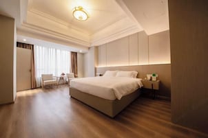 Quarto de hotel com cama grande, mesa de cabeceira, cadeira e janela com cortinas.