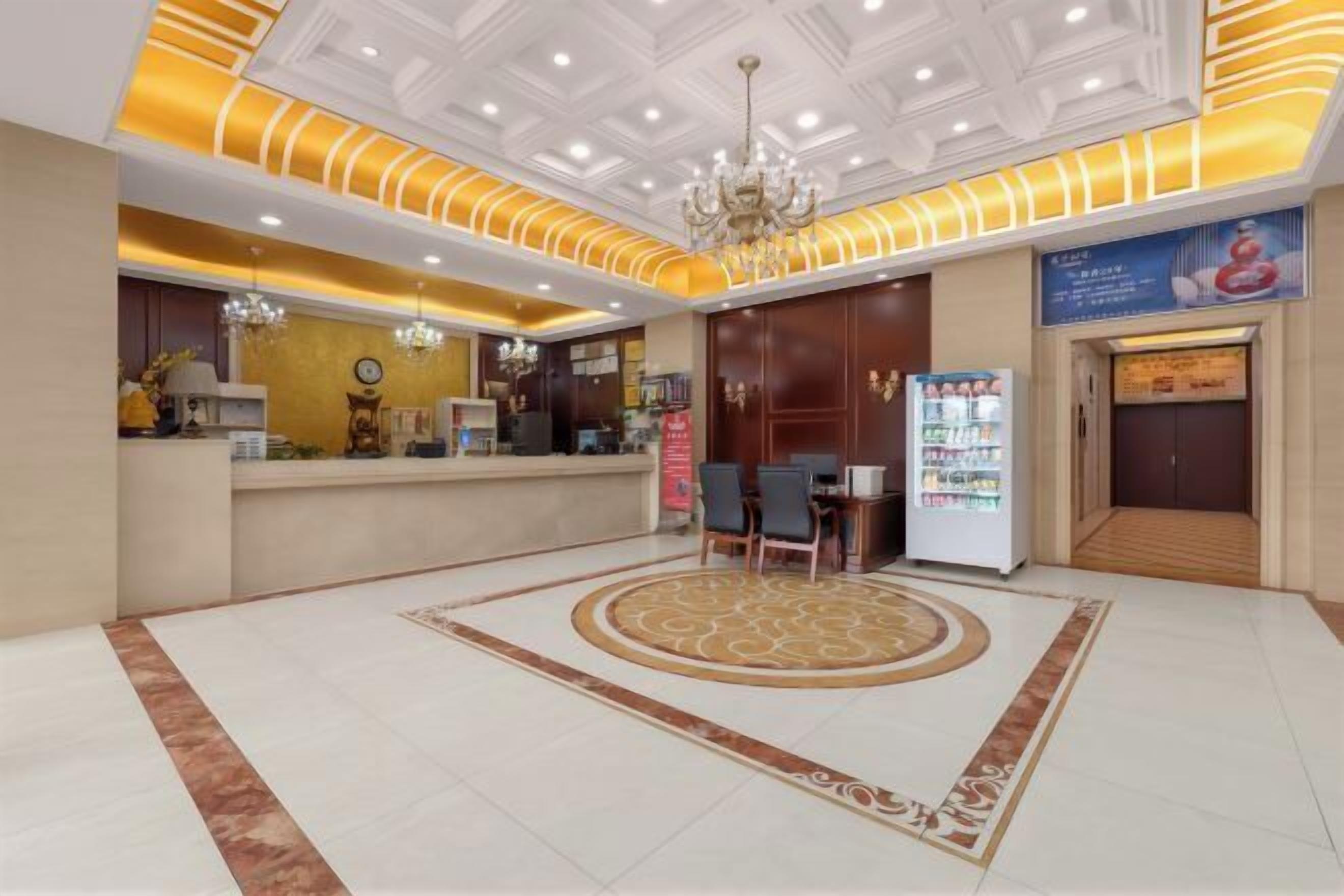 Um saguão de hotel com uma mesa de recepção, um candelabro, uma máquina de vendas automática e um tapete estampado.