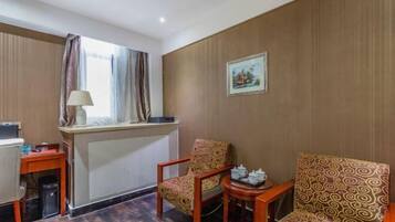 Quarto de hotel com escrivaninha, duas cadeiras estampadas, uma mesa pequena com chaleiras, janela com cortinas e um quadro emoldurado na parede.