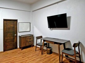 Quarto com piso de madeira, escrivaninha com duas cadeiras, televisão fixada na parede, armário de madeira e uma porta.