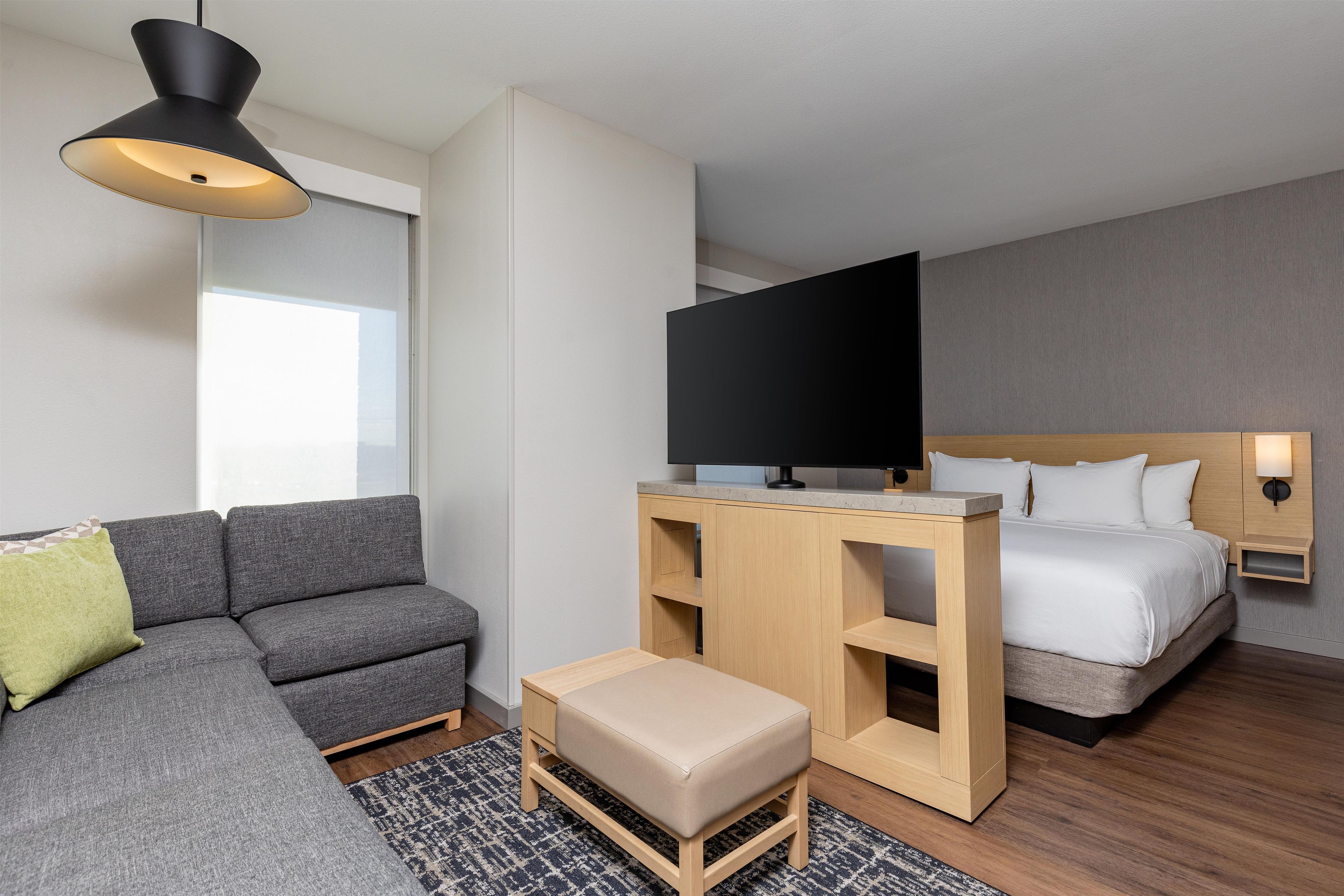 Quarto de hotel moderno com sofá cinza, escrivaninha de madeira com TV de tela plana, cama com roupa de cama branca e uma janela grande que oferece luz natural.