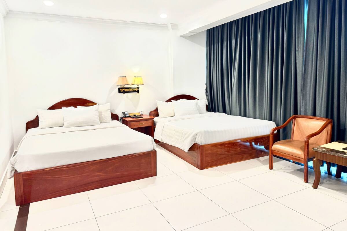 Quarto de hotel com duas camas de madeira, uma mesa de cabeceira com abajur, uma cadeira e uma janela grande com cortinas escuras.
