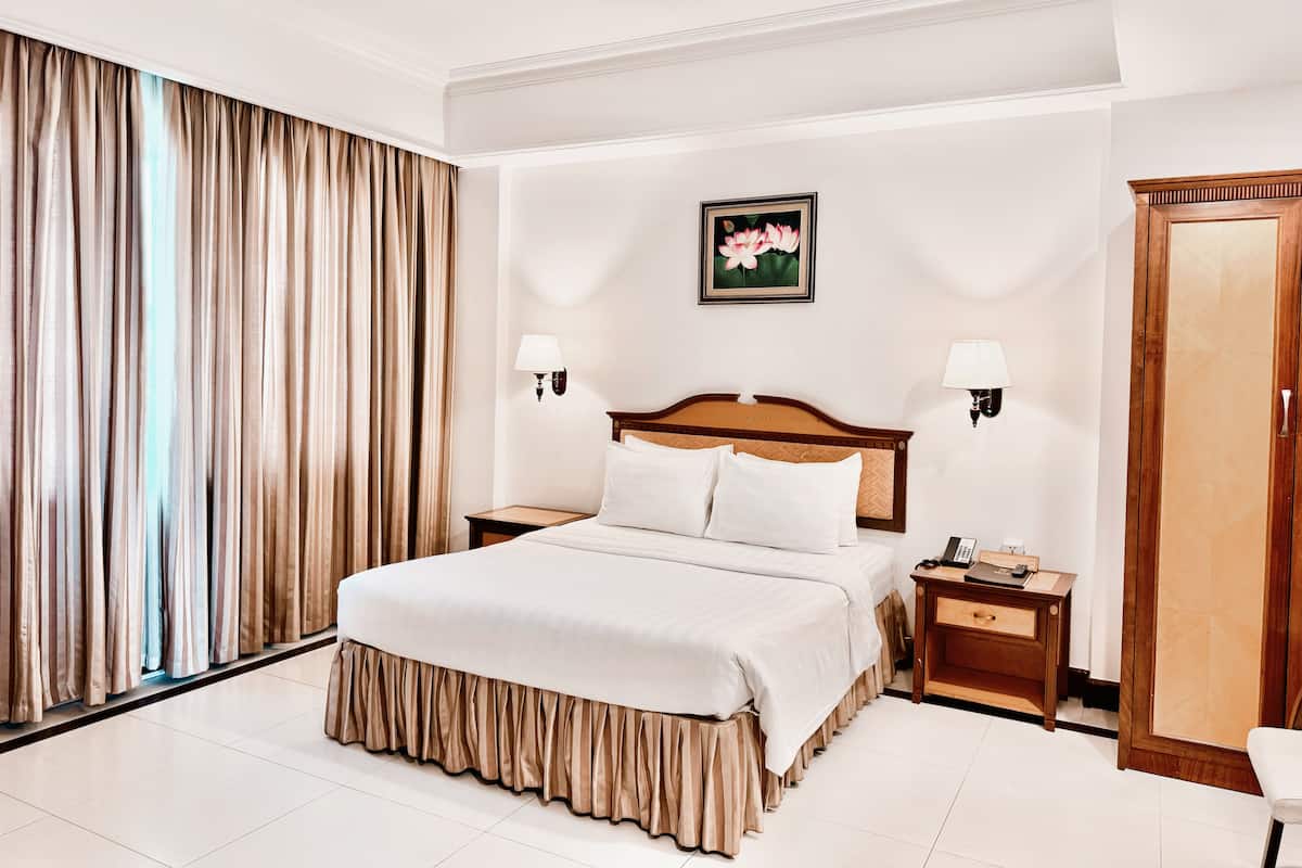 Quarto de hotel com uma cama grande, duas mesas de cabeceira, um criado-mudo, um guarda-roupa de madeira e uma janela ampla com cortinas.