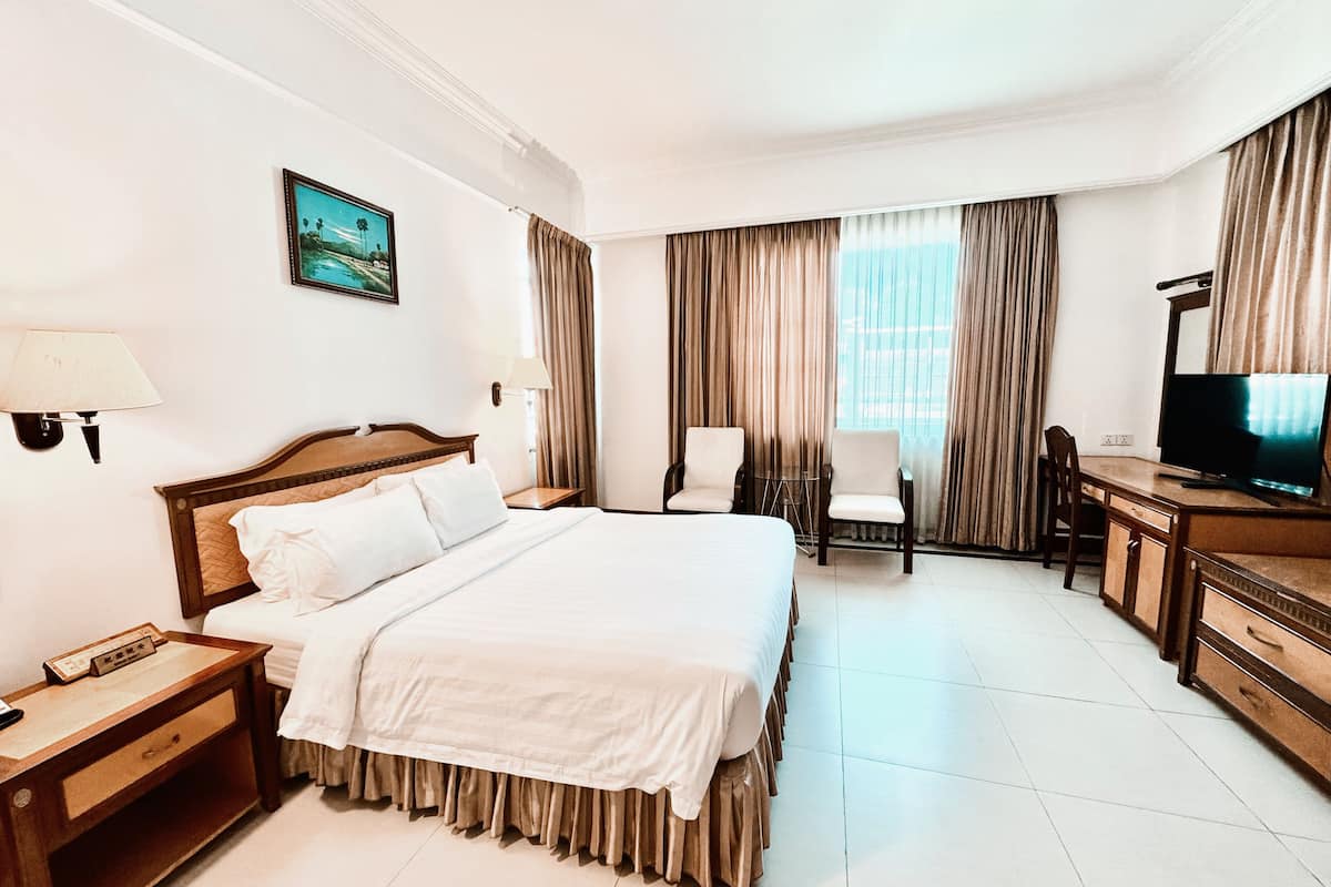 Quarto de hotel com uma cama grande, uma escrivaninha com televisão, duas cadeiras e uma janela com cortinas.