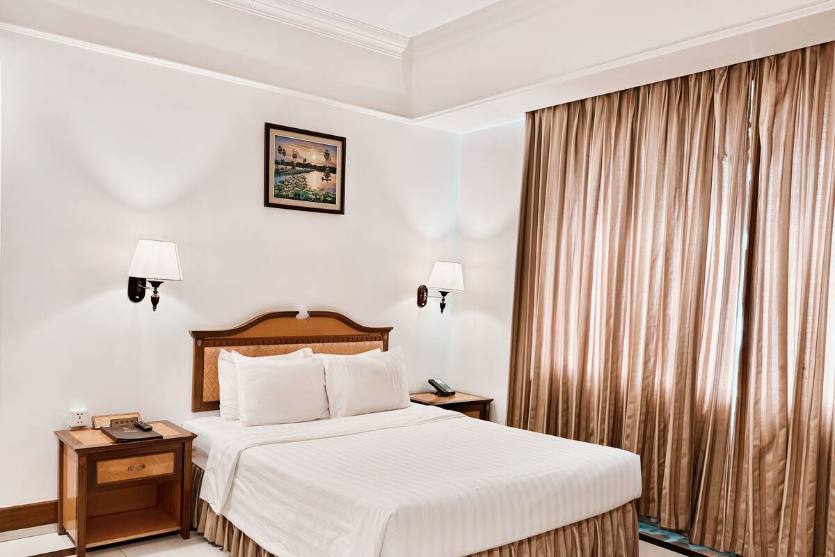 Quarto de hotel com uma cama grande, duas mesas de cabeceira, um criado-mudo, um quadro na parede e uma cortina.