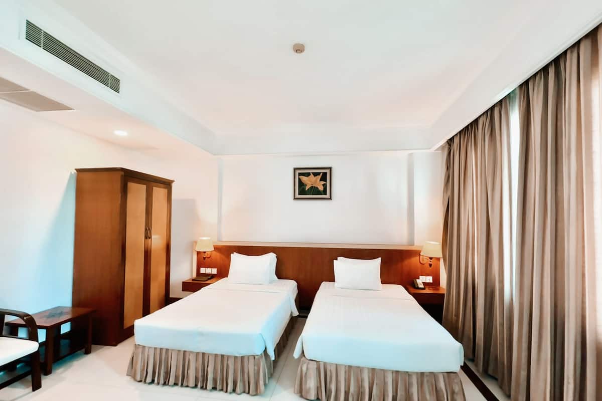 Quarto de hotel com duas camas de solteiro, um guarda-roupa de madeira, uma escrivaninha e uma cadeira.