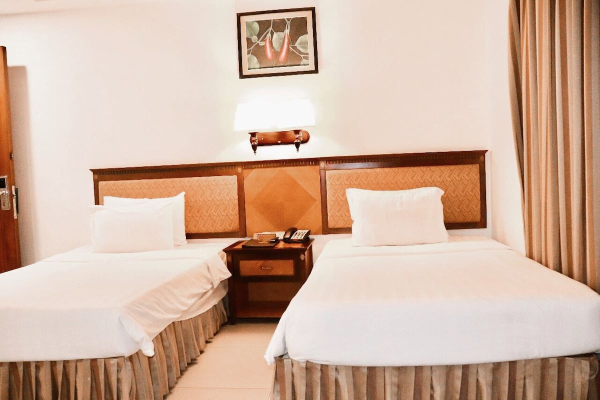 Quarto de hotel com duas camas de solteiro, mesa de cabeceira com abajur, telefone e um quadro na parede.