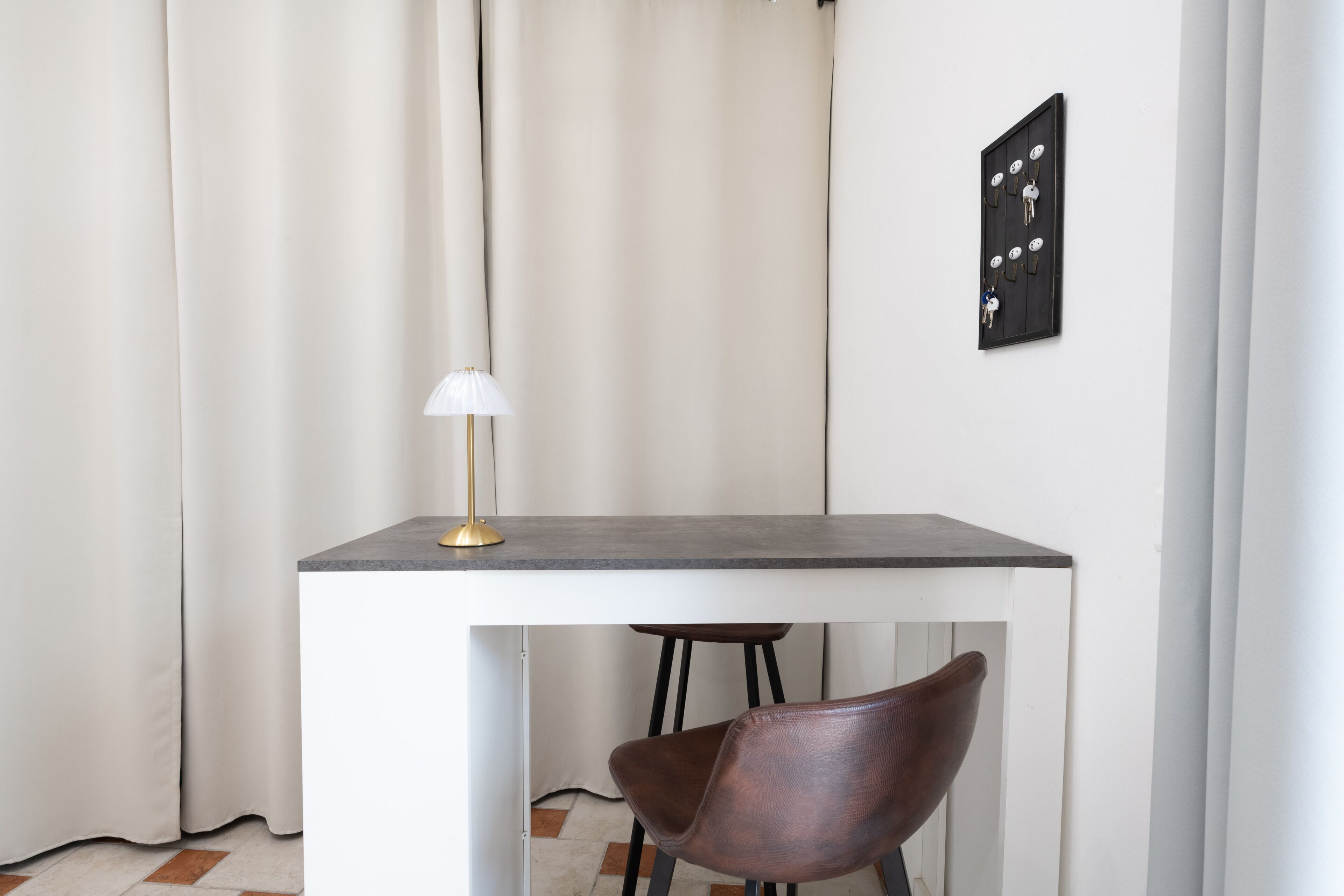 Quarto pequeno e moderno com uma mesa branca, uma cadeira marrom, um abajur e um interruptor de luz na parede.