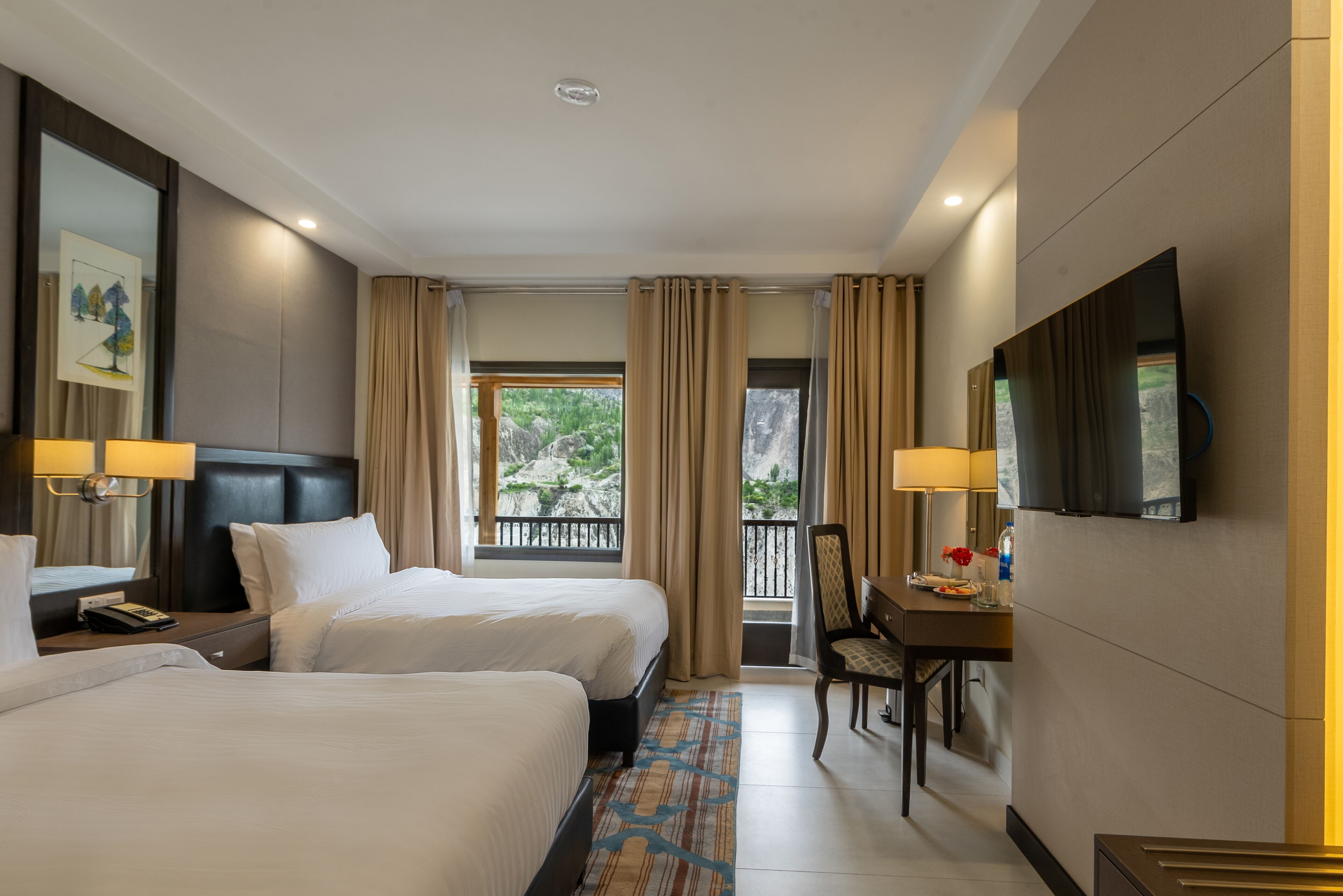 Quarto de hotel com duas camas, uma escrivaninha, uma cadeira, uma televisão e uma varanda com cortinas.