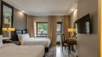 Quarto de hotel com duas camas, uma escrivaninha, uma cadeira, uma televisão e uma varanda com cortinas.