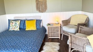 Quarto com cama, uma cadeira, mesa pequena com abajur e um enfeite de macramê na parede.