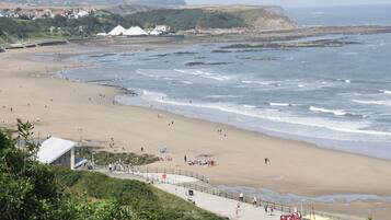 Uma praia costeira com um calçadão pavimentado, uma torre salva-vidas e uma vista distante de uma cidade.