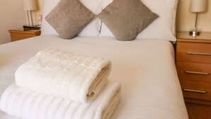 Cama bem arrumada com toalha branca dobrada, duas almofadas e cabeceira de madeira.