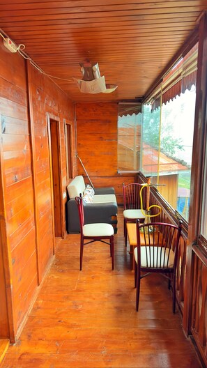 Interior de uma cabana de madeira com sofá, cadeiras, mesa de jantar e janela com vista para uma casa.