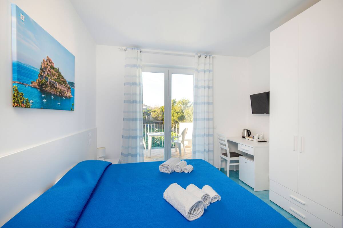 Quarto de hotel com cama, escrivaninha, televisão e varanda com vista para o mar.