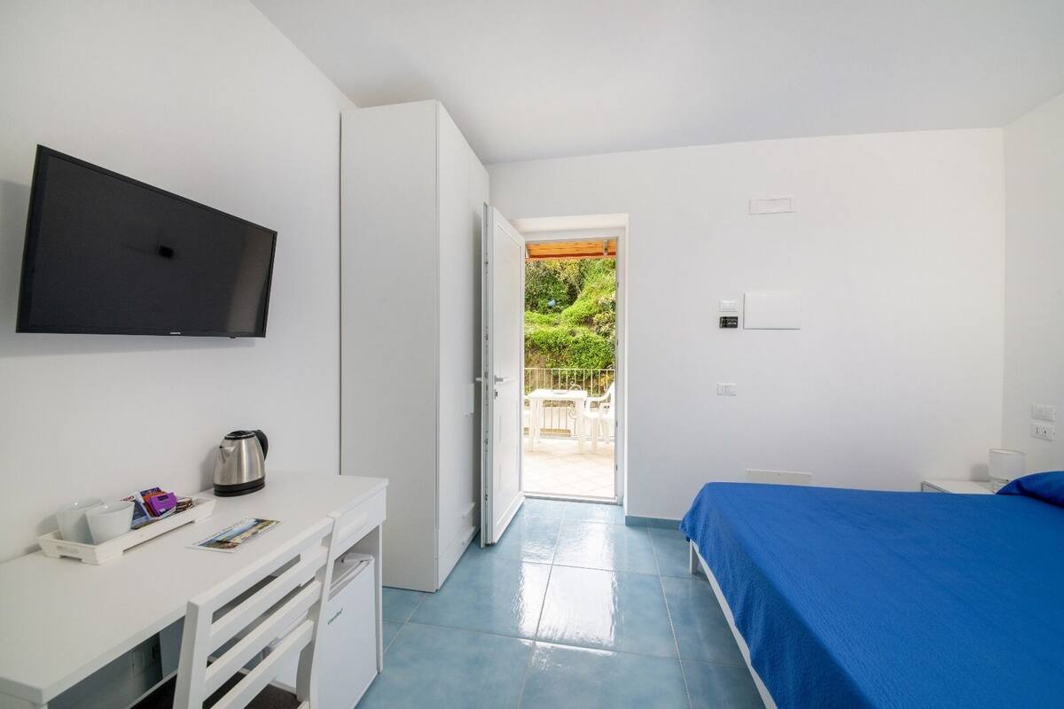 Quarto com cama, televisão, escrivaninha com chaleira e vista para um jardim através de uma porta aberta.