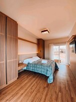 Quarto moderno com piso de madeira, cama com colcha azul, mesa de cabeceira e uma janela grande com cortinas.
