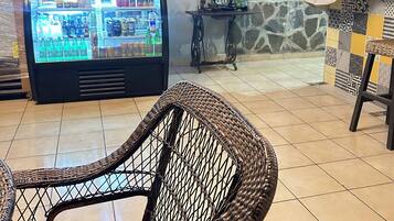Um lobby de hotel com balcão de recepção, refrigerador exibindo bebidas, cadeiras de vime e um painel decorado com árvore de Natal e luzes.