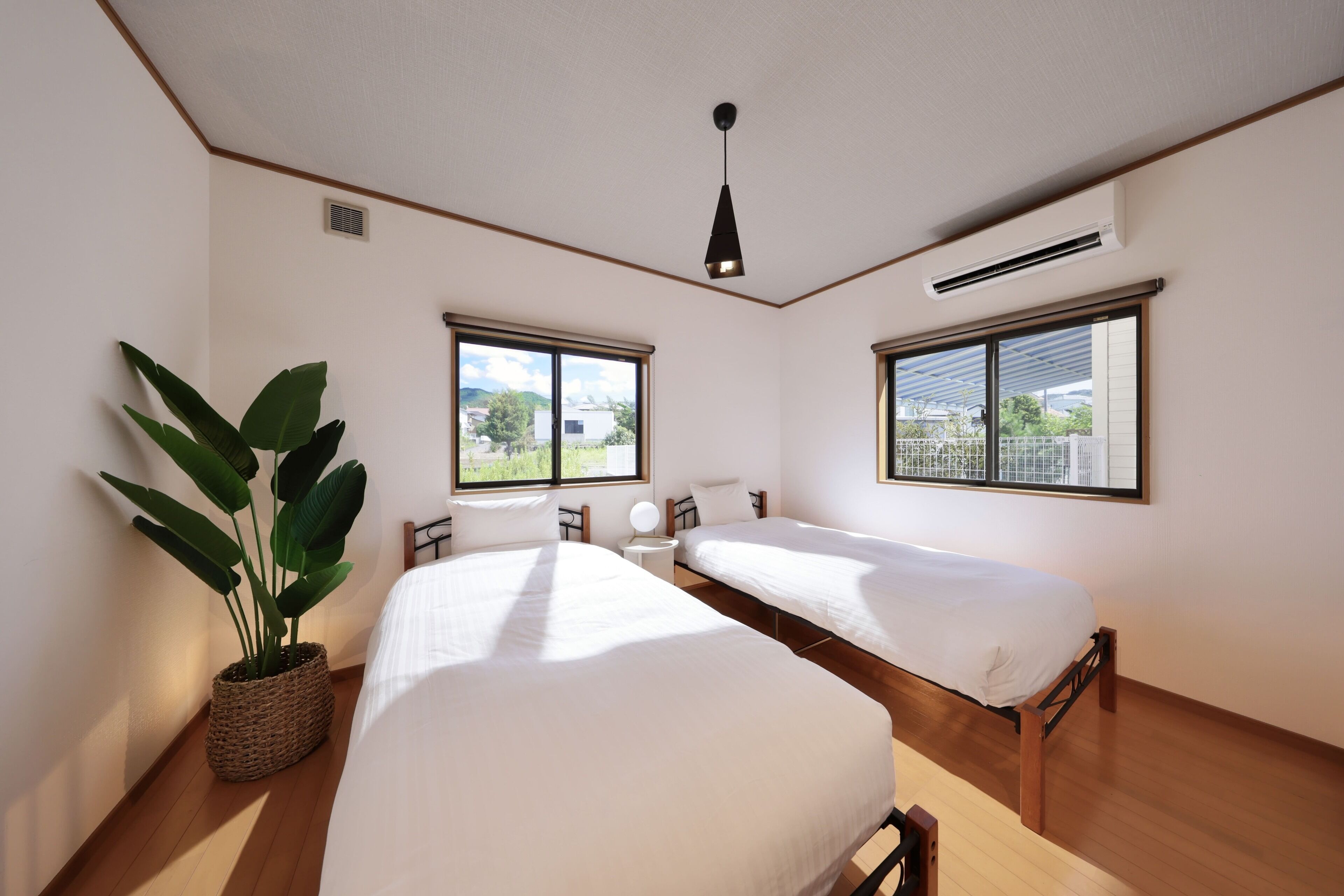 Quarto com duas camas, janelas amplas, ventilador de teto e um vaso com planta.