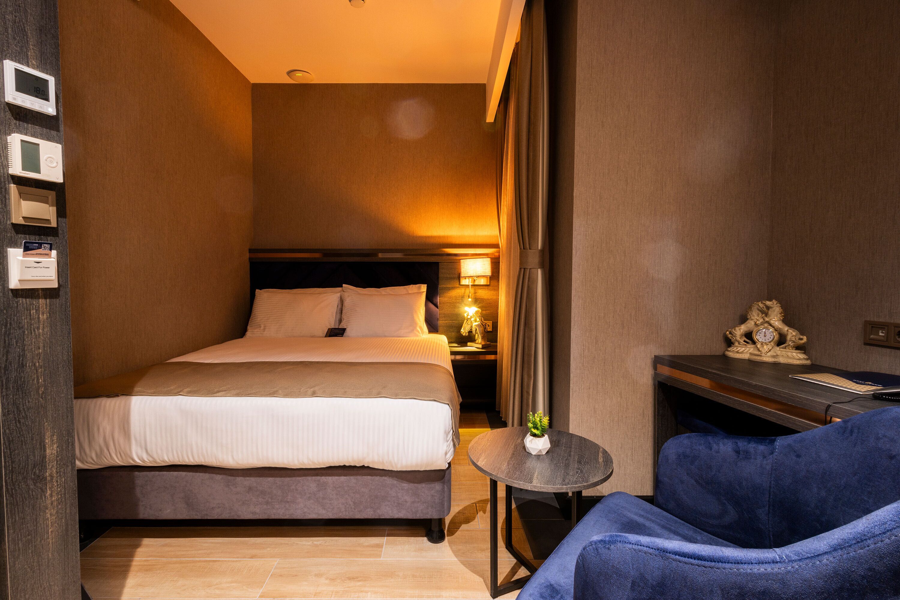 Quarto de hotel com uma cama, uma mesinha de cabeceira com abajur, uma cadeira e uma escrivaninha com computador.