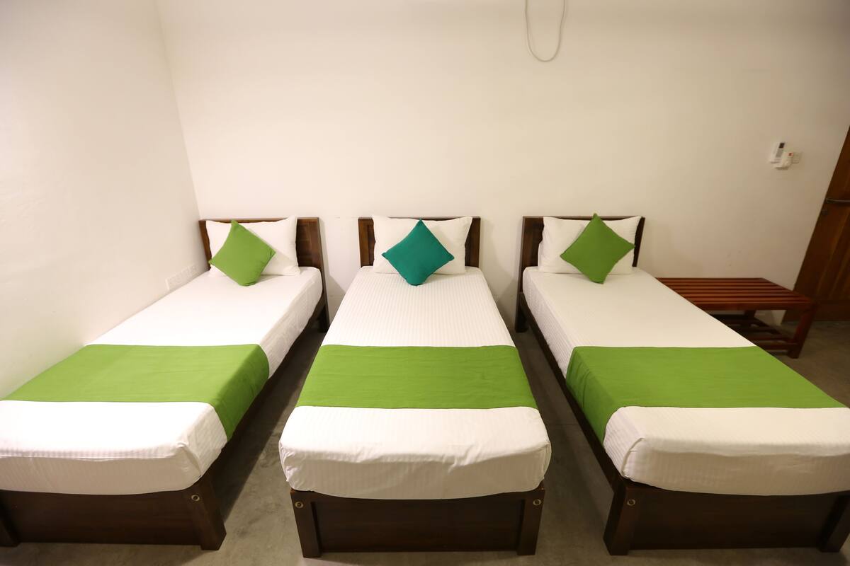 Três camas de solteiro com roupa de cama verde e branca, cabeceiras de madeira e um pequeno banco de madeira.