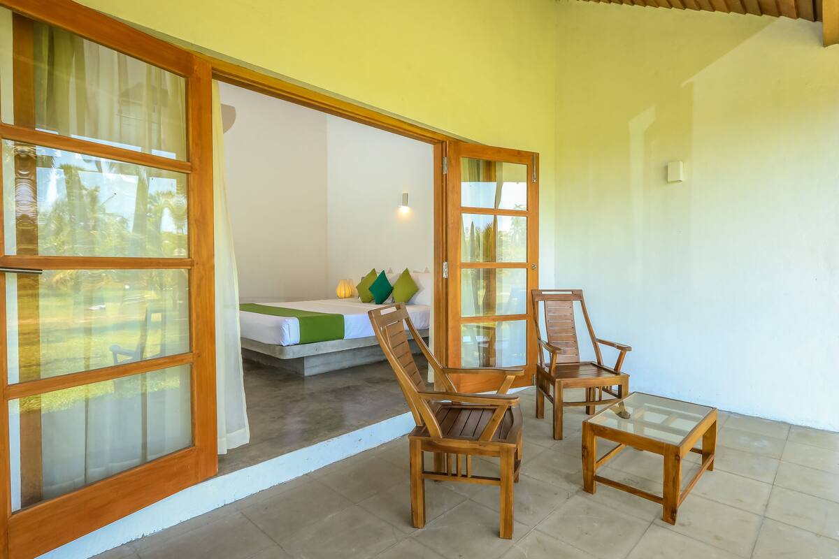 Um quarto com uma cama, duas cadeiras de madeira, uma mesa de vidro e grandes janelas com cortinas.