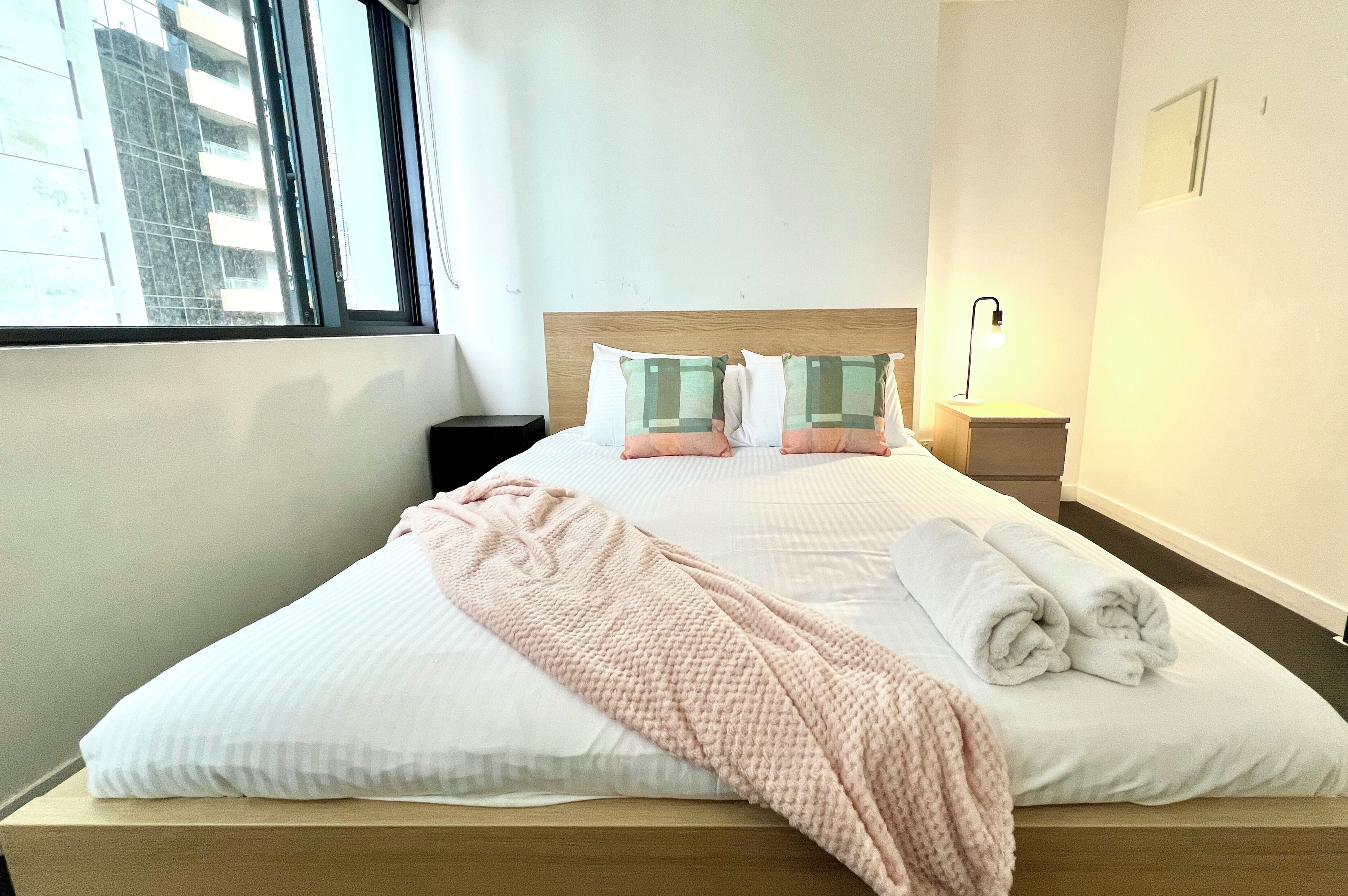 Cama bem arrumada com almofadas e cobertor, mesa de cabeceira com luminária e janela com vista para edifícios.