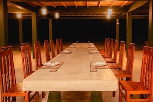 Uma longa mesa de jantar com cadeiras, posta para uma refeição, em área externa coberta com vigas de madeira e luminárias pendentes.